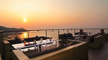 Terraço no terraço com móveis de vime e uma mesa posta para refeições, com vista para um pôr do sol sobre a água.