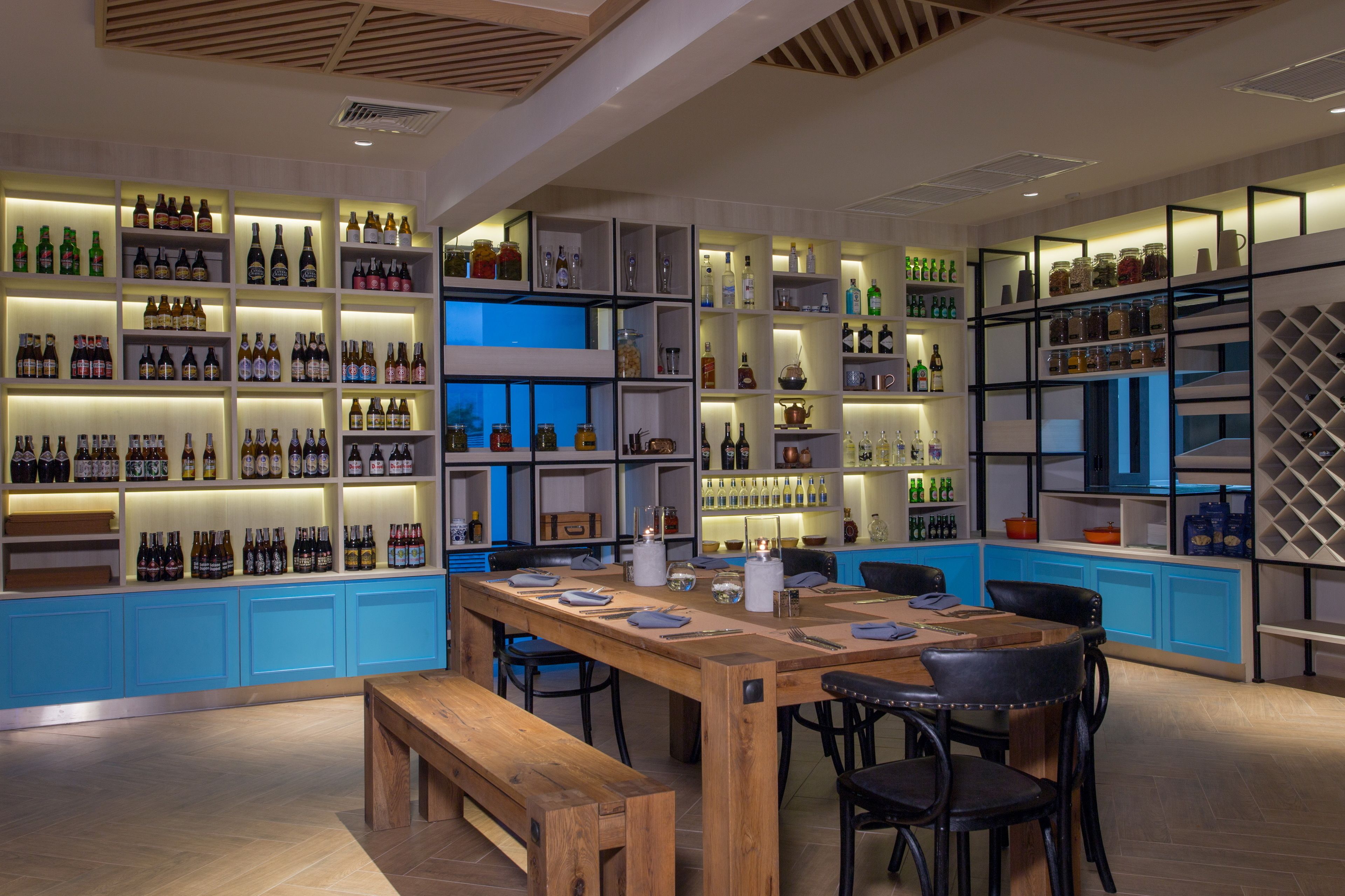 Uma área de bar com prateleiras de garrafas e copos, uma mesa de madeira com cadeiras e um banco.