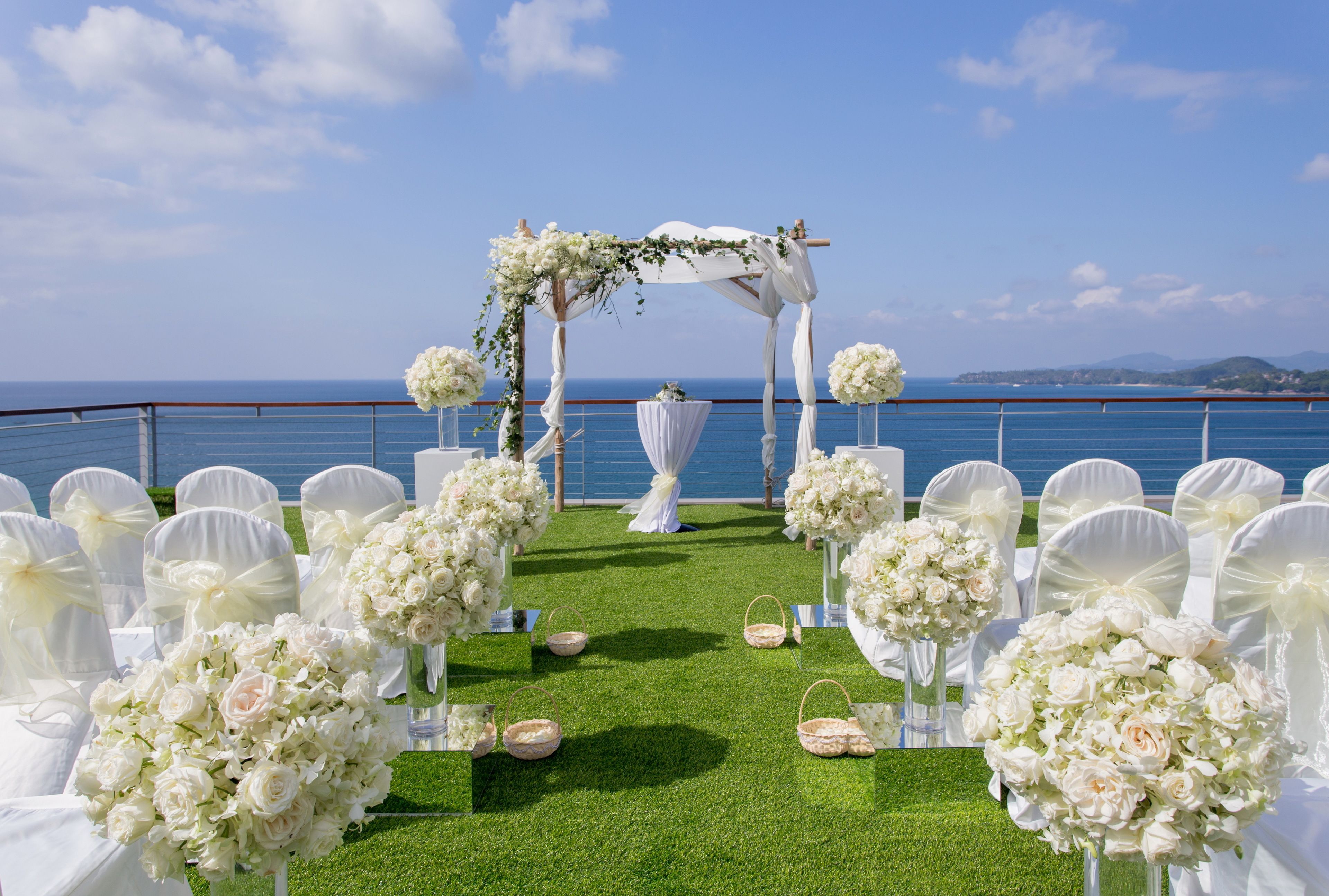 Preparação para cerimônia de casamento com um arco branco e arranjos florais em uma área gramada perto do mar.
