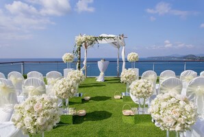 Preparação para cerimônia de casamento com um arco branco e arranjos florais em uma área gramada perto do mar.