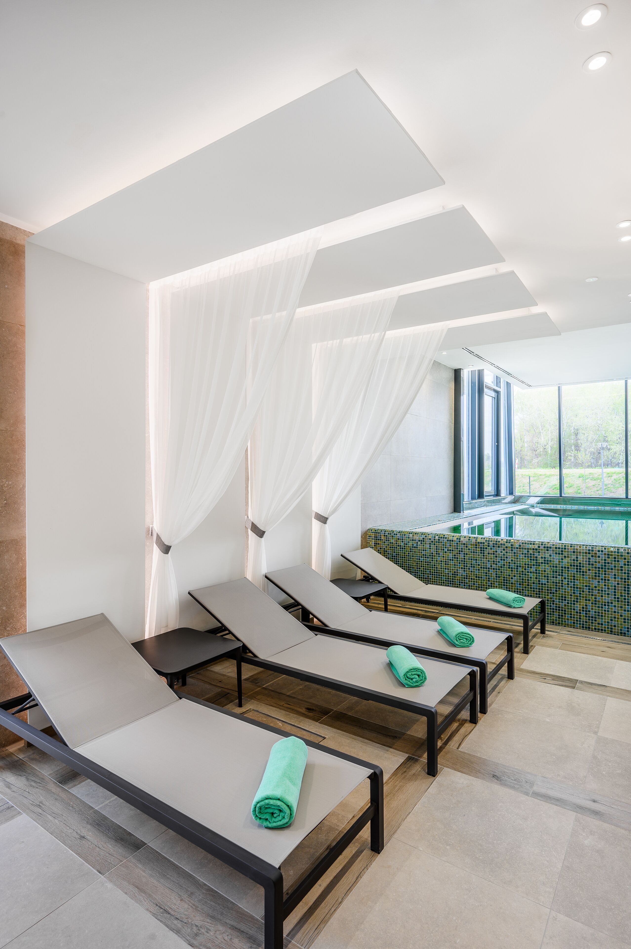 Um salão de spa com cadeiras reclináveis, uma piscina em vidro e vista para o verde lá fora.