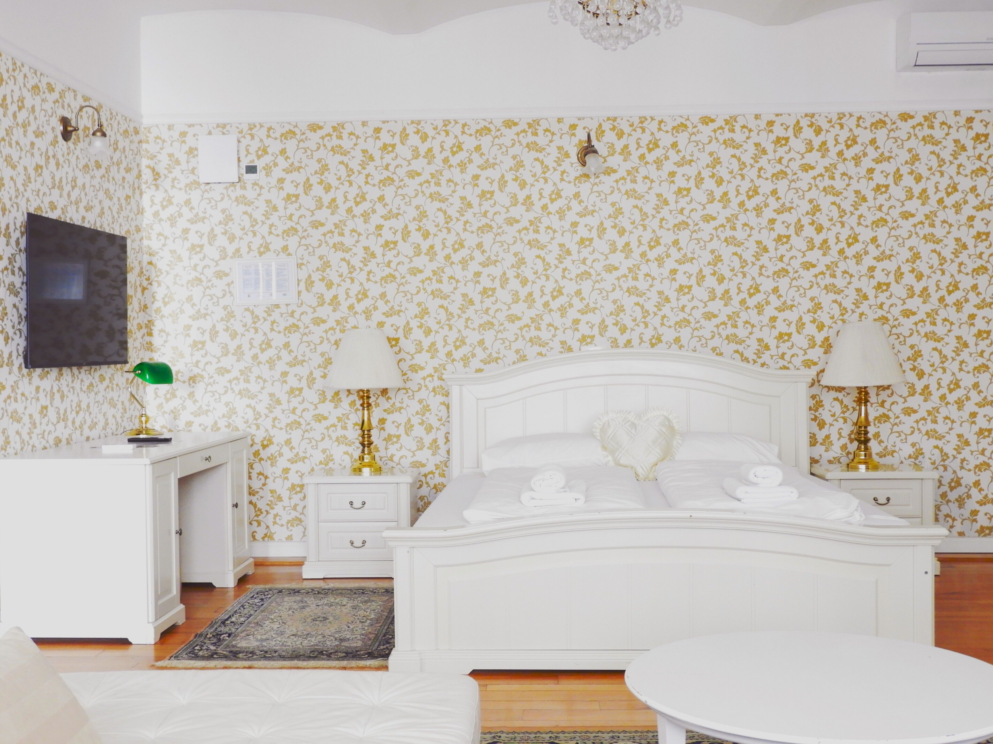 Quarto com cama branca, duas mesas de cabeceira com luminárias, escrivaninha com abajur, mesinha redonda e papel de parede estampado.