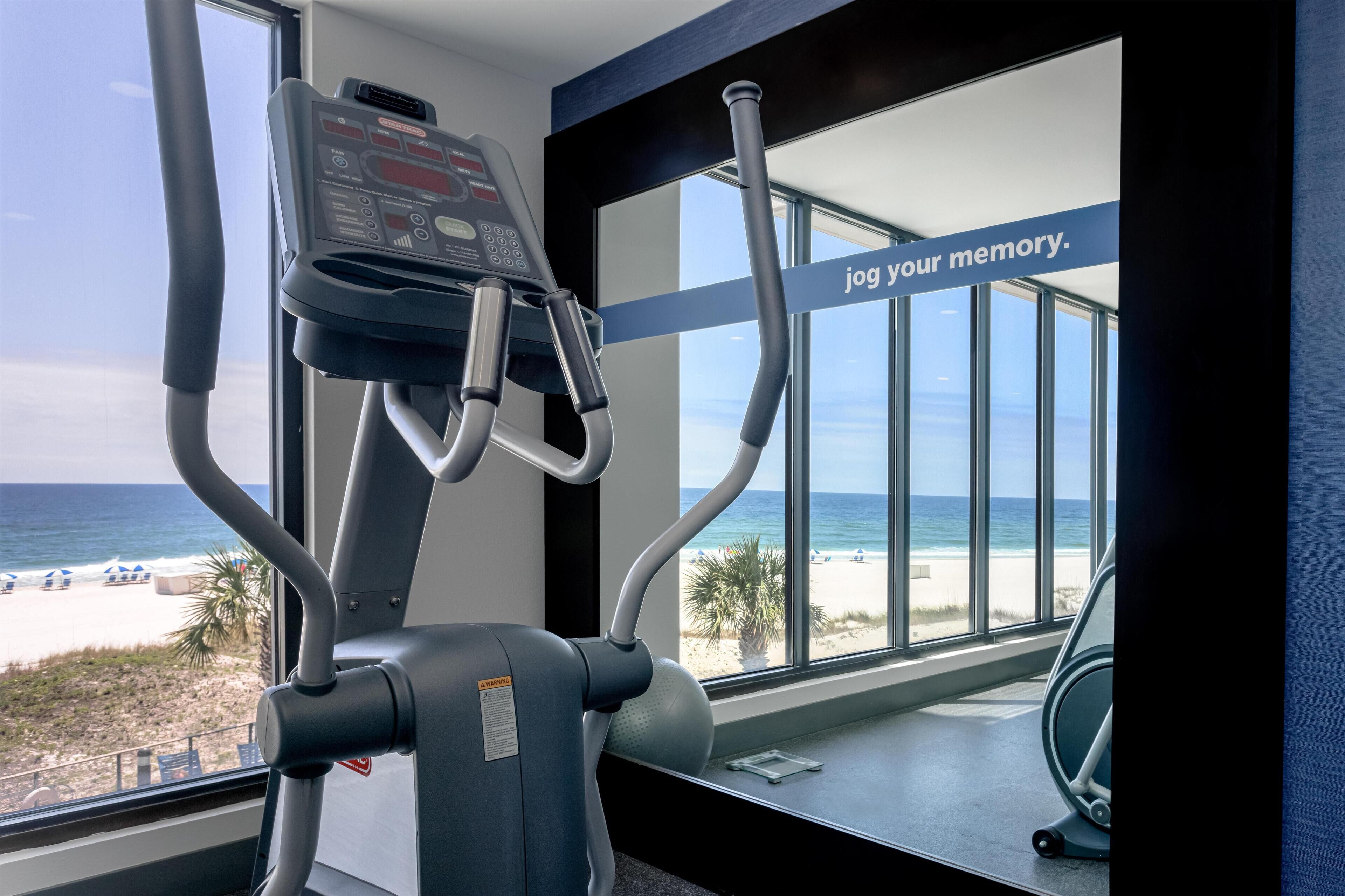 Uma academia com esteira e equipamentos de exercícios, com vista para uma praia com palmeiras e céu claro.