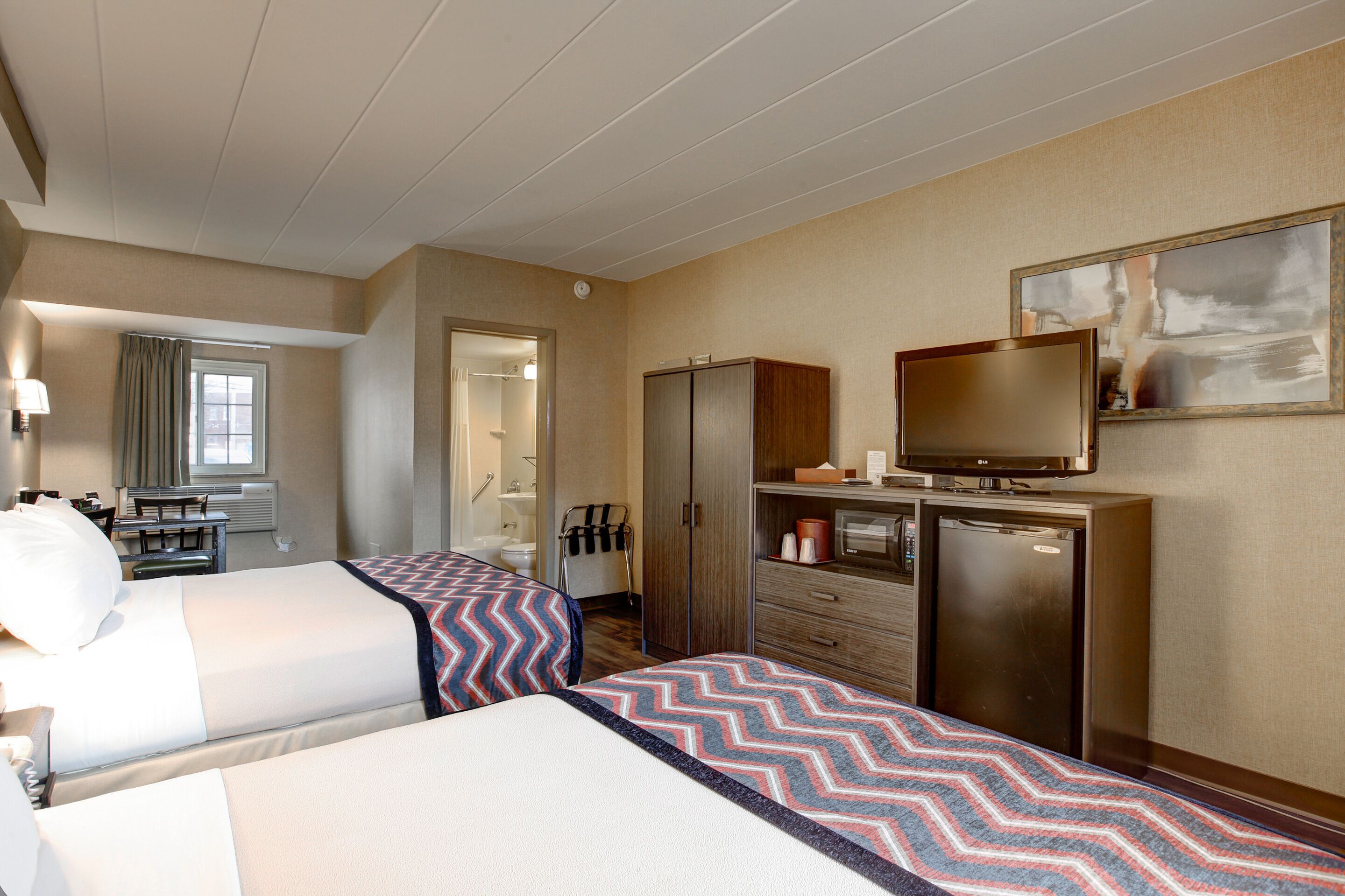 Quarto de hotel com duas camas, uma escrivaninha, televisão, guarda-roupa e casa de banho visível através de uma porta aberta.