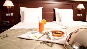 Quarto de hotel com uma cama bem arrumada, bandeja de café da manhã com torradas, manteiga e um copo de suco de laranja, e uma mesa de cabeceira com abajur e relógio.