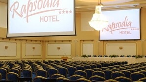 Uma sala de conferências no Hotel Rapsodia, com fileiras de cadeiras azuis, um pódio e uma tela grande exibindo o nome do hotel.