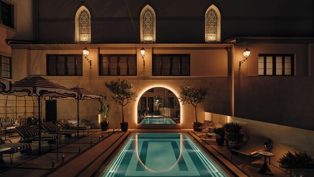 Uma área de piscina com espreguiçadeiras e guarda-sóis, uma fonte e um edifício com janelas em arco, além de um espaço para sentar à beira da piscina.