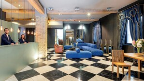 Um saguão de hotel com piso xadrez, uma mesa de recepção, um sofá azul e uma mesa de jantar de madeira.