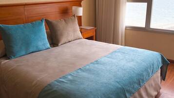 Uma cama com cabeceira de madeira, uma mesa de cabeceira com abajur e vista para o mar pela janela.