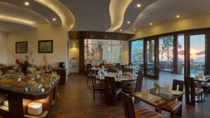 Um restaurante com buffet à esquerda, mesas e cadeiras para refeições ao centro, e uma grande janela com vista para o exterior.