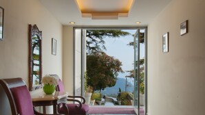 Um corredor com porta de vidro que dá para uma varanda com vista para o mar. Há duas cadeiras e uma mesa com um vaso de flores.