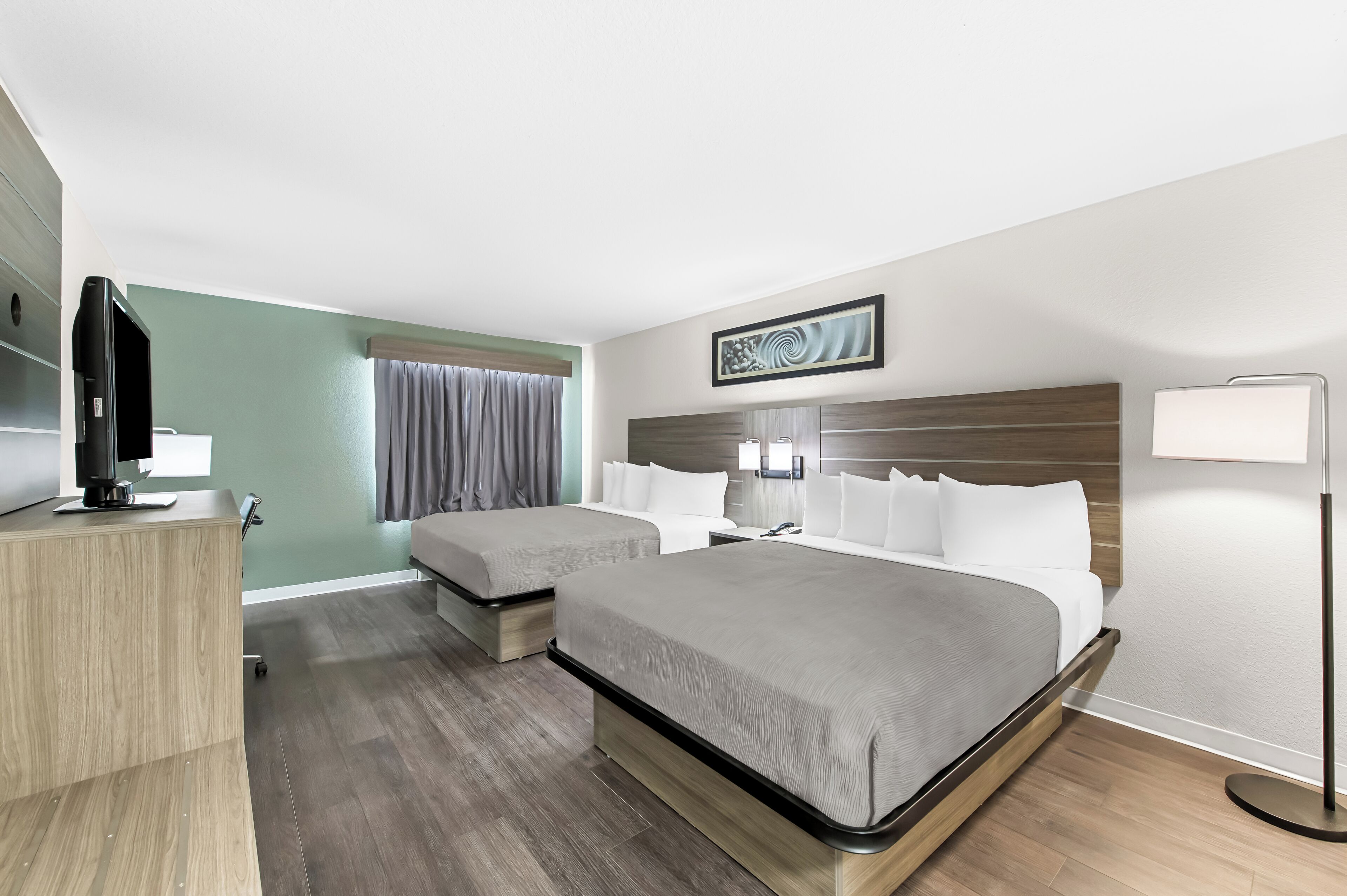 Quarto de hotel com duas camas, uma mesa de escritório com televisão, mesa de cabeceira, abajur e janela com cortinas.