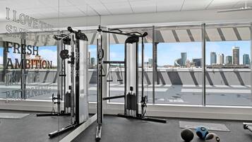 Uma academia com equipamentos de ginástica, uma frase motivacional na parede e vista para a paisagem urbana através de grandes janelas.