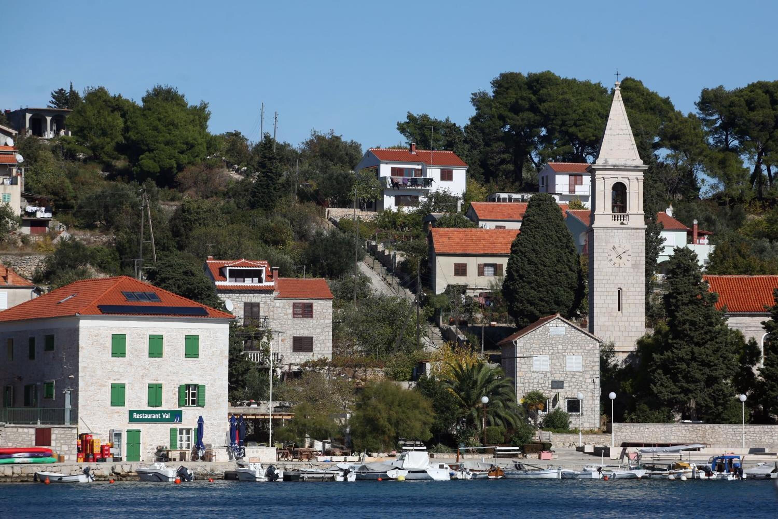 Uma cidade costeira com uma torre de igreja imponente, um restaurante chamado 'Restaurant Val' e várias embarcações no porto.