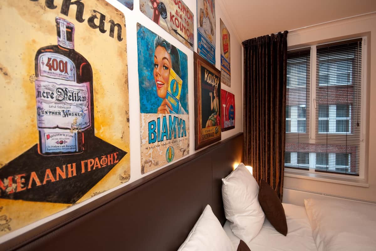 Quarto de hotel com uma cama, almofadas, uma janela com persianas e uma parede decorada com cartazes antigos.
