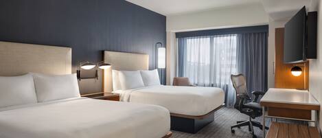 Quarto de hotel com duas camas, uma escrivaninha, uma cadeira, televisão e janela com cortinas.