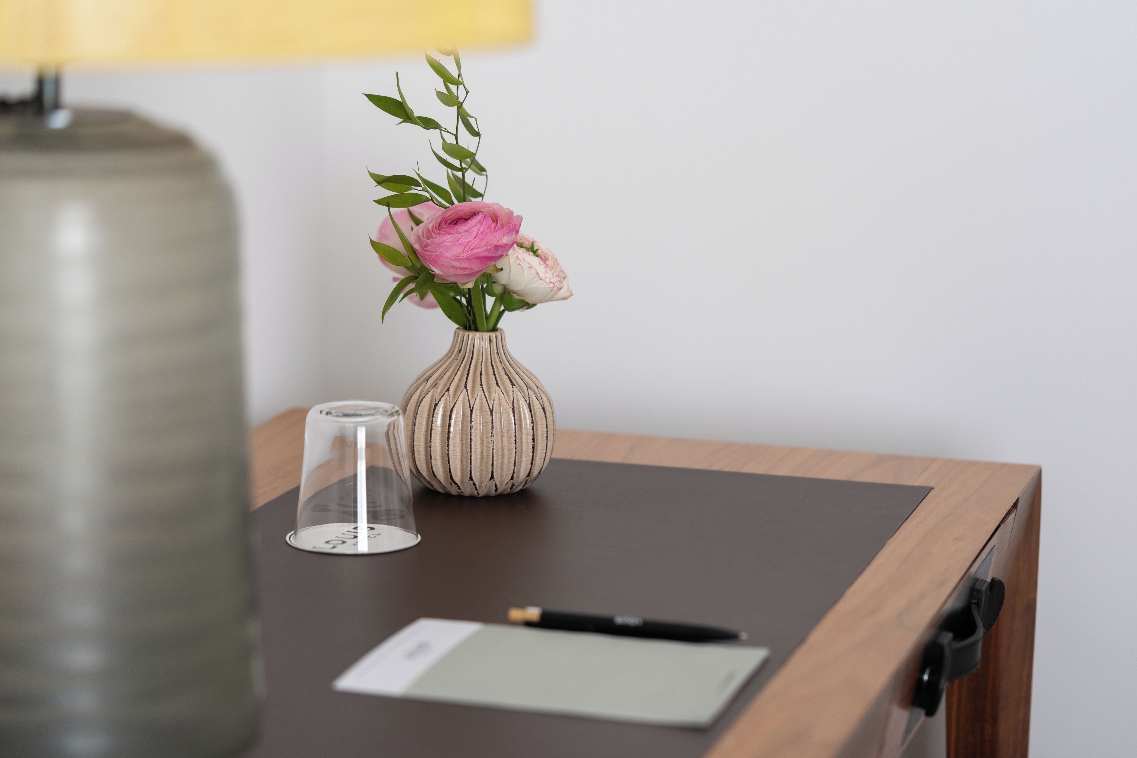 Uma mesa com um vaso de flores, um copo de vidro transparente e uma caneta preta sobre um bloco de notas.