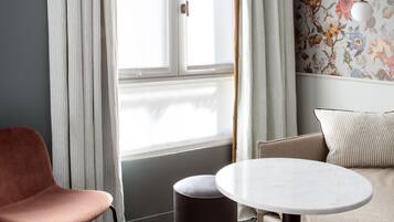Quarto moderno com sofá, mesa redonda e uma cadeira. Há uma janela com cortinas, parede estampada e luminária pendente.