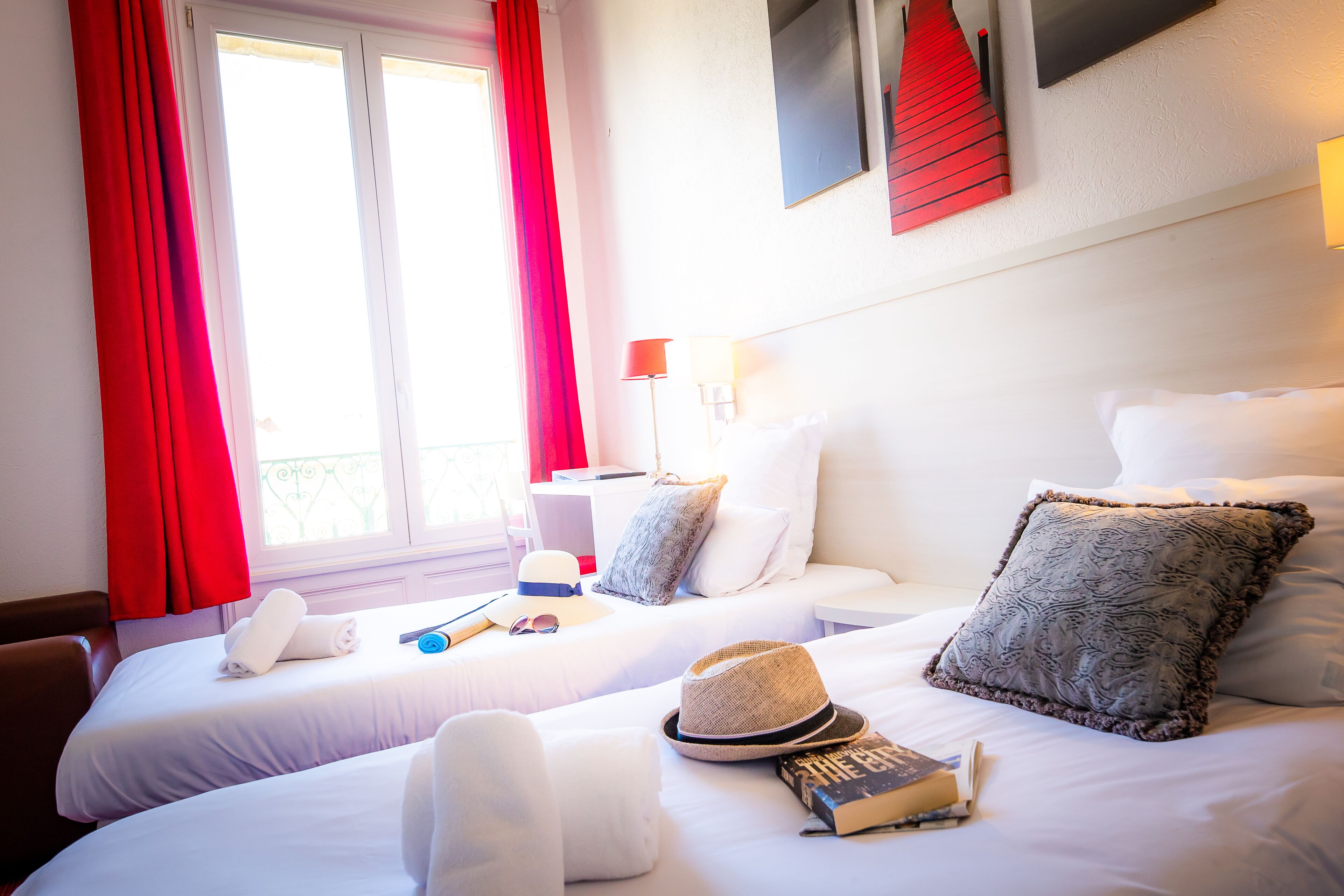 Quarto de hotel com duas camas, janela com cortinas vermelhas, televisão na parede e mesa de cabeceira com abajur.