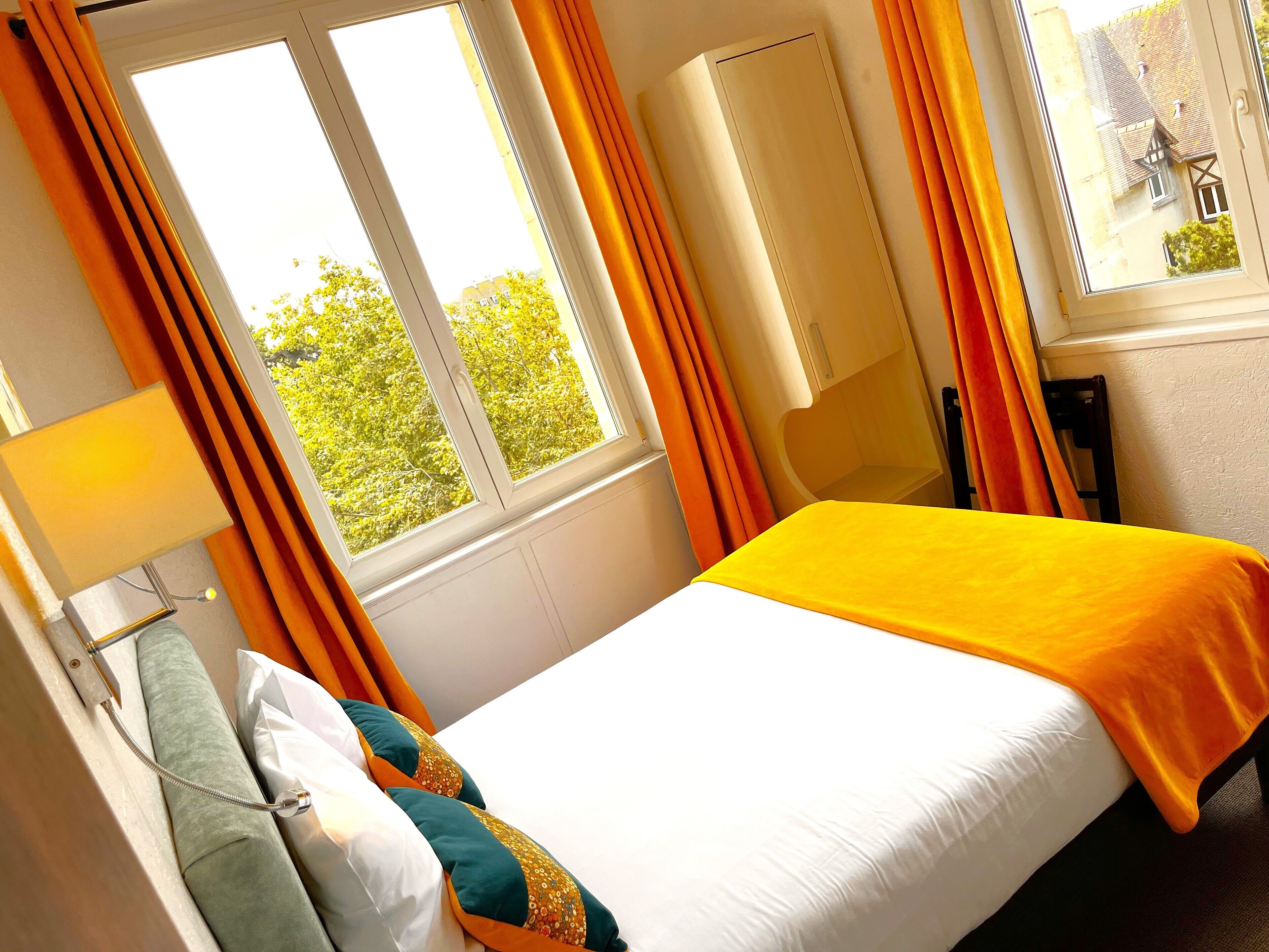 Quarto de hotel com cama, janela com cortinas, abajur e vista para o verde lá fora.