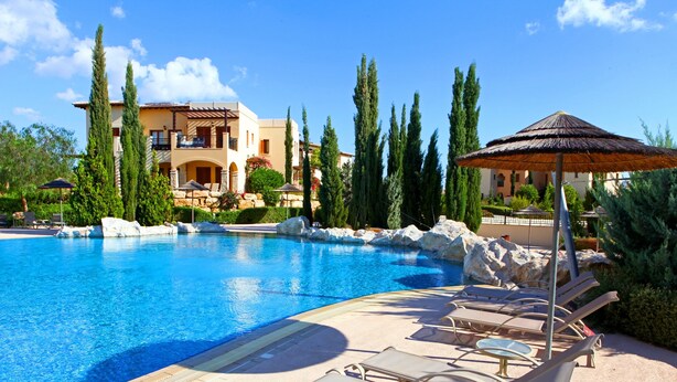 Piscina com espreguiçadeiras e guarda-sóis, cercada por árvores altas e um edifício com sacadas.