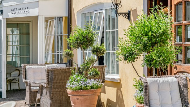 Móveis de vime, plantas em vasos e um edifício com a placa 'LATE ARRIVAL NATINDGANG'.