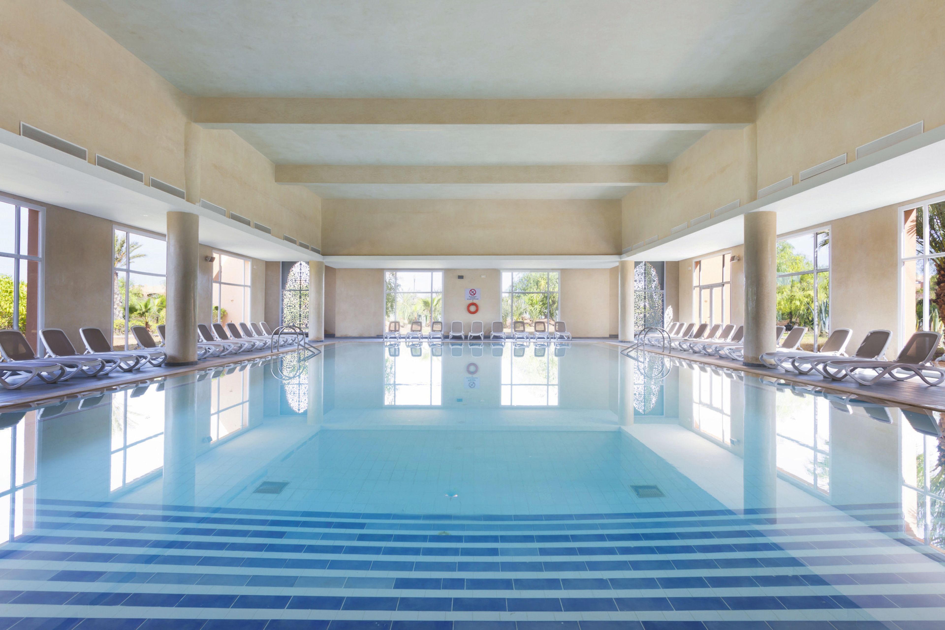 Uma piscina interior ampla com água azul cristalina, rodeada por espreguiçadeiras e grandes janelas.