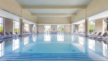 Uma piscina interior ampla com água azul cristalina, rodeada por espreguiçadeiras e grandes janelas.