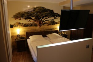 Quarto de hotel com uma cama, mesa de cabeceira, abajur, telefone e televisão na parede.
