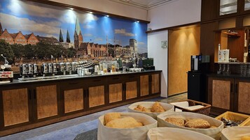 Um saguão de hotel com um grande mural da paisagem urbana, um café com diversas máquinas e recipientes de café, e áreas de estar com cestas de pão.