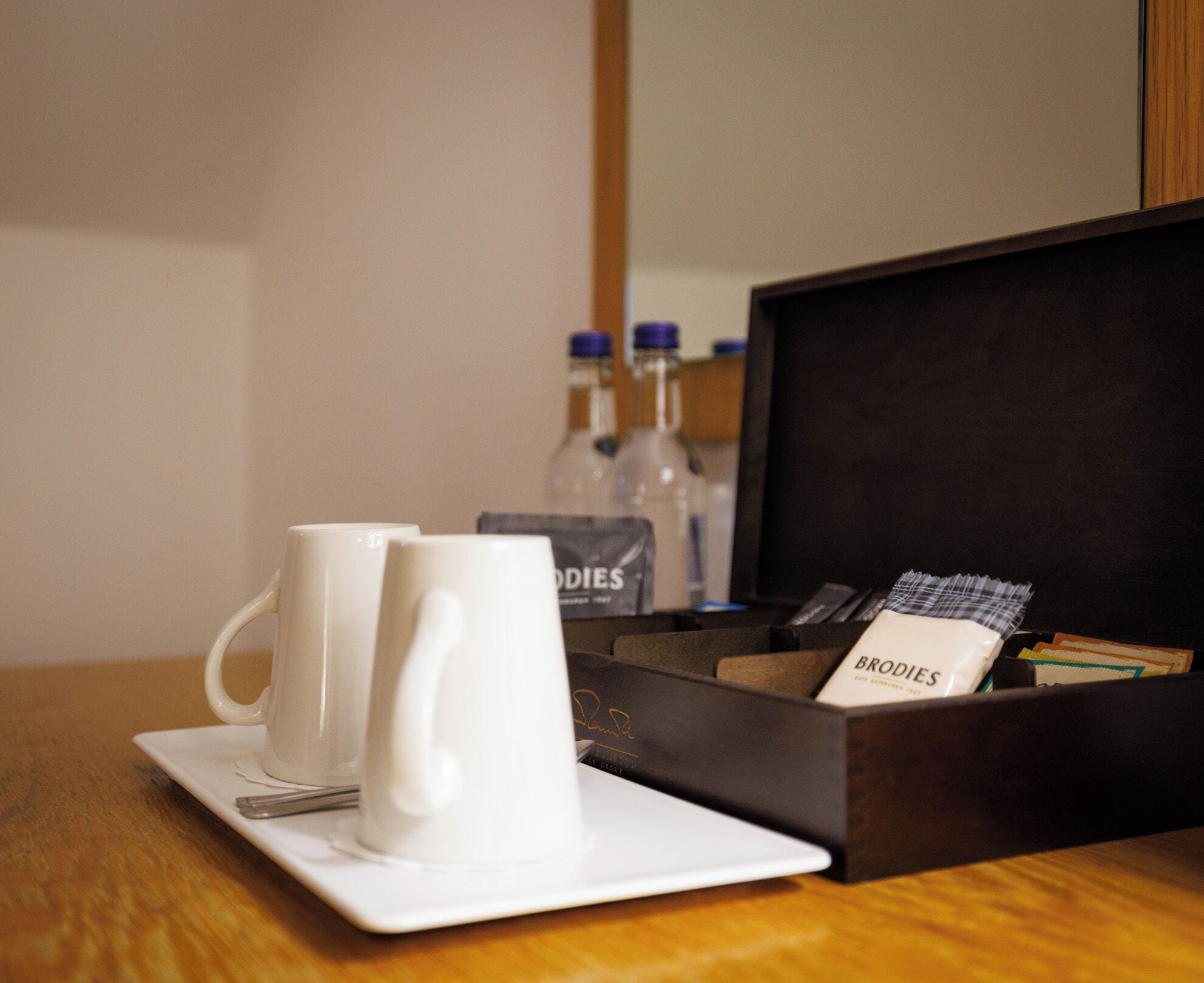 Quarto de hotel com uma mesa de madeira, duas canecas brancas, um prato e uma caixa com artigos de higiene pessoal.