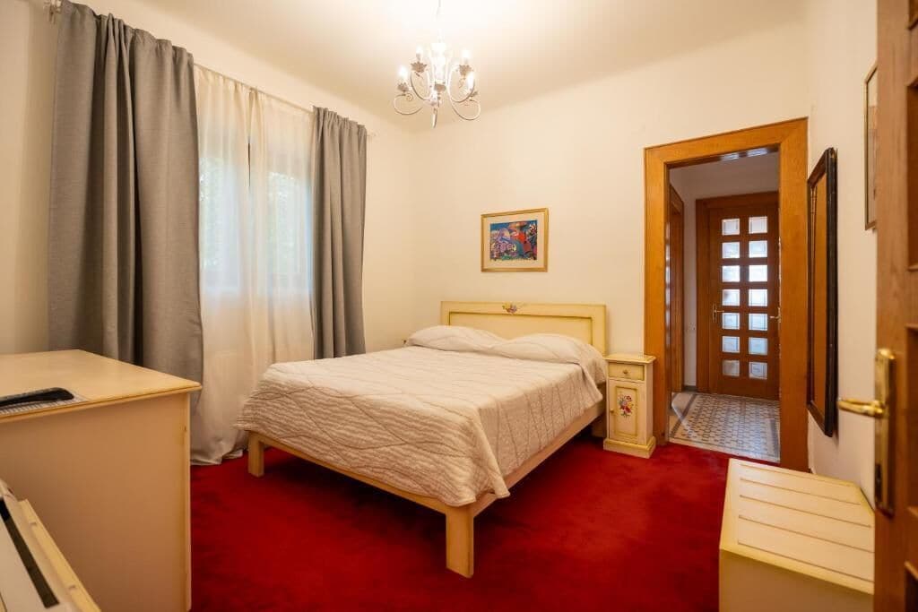 Um quarto com cama, escrivaninha, penteadeira, lustre, porta e um quadro na parede.