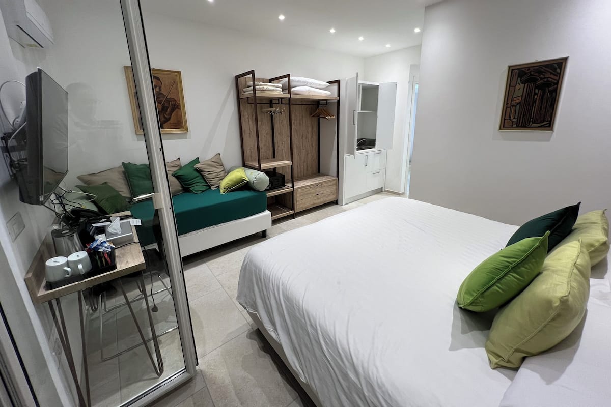 Quarto moderno com cama, área de estar com almofadas, guarda-roupa e televisão.