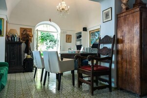 Uma sala de jantar com um candelabro, uma cadeira de madeira com almofada vermelha, um sofá verde e um armário.