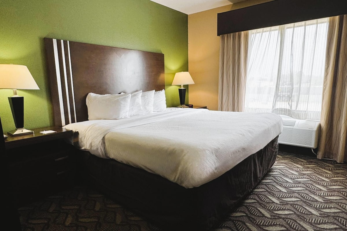 Quarto de hotel com uma cama grande, duas mesas de cabeceira com abajures, uma janela com cortinas e um tapete estampado.