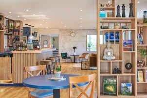 Uma área de bar moderna com balcão, banquetas e diversos tipos de garrafas. Há também uma área de estar com mesa e cadeiras, e uma estante com livros e objetos decorativos.