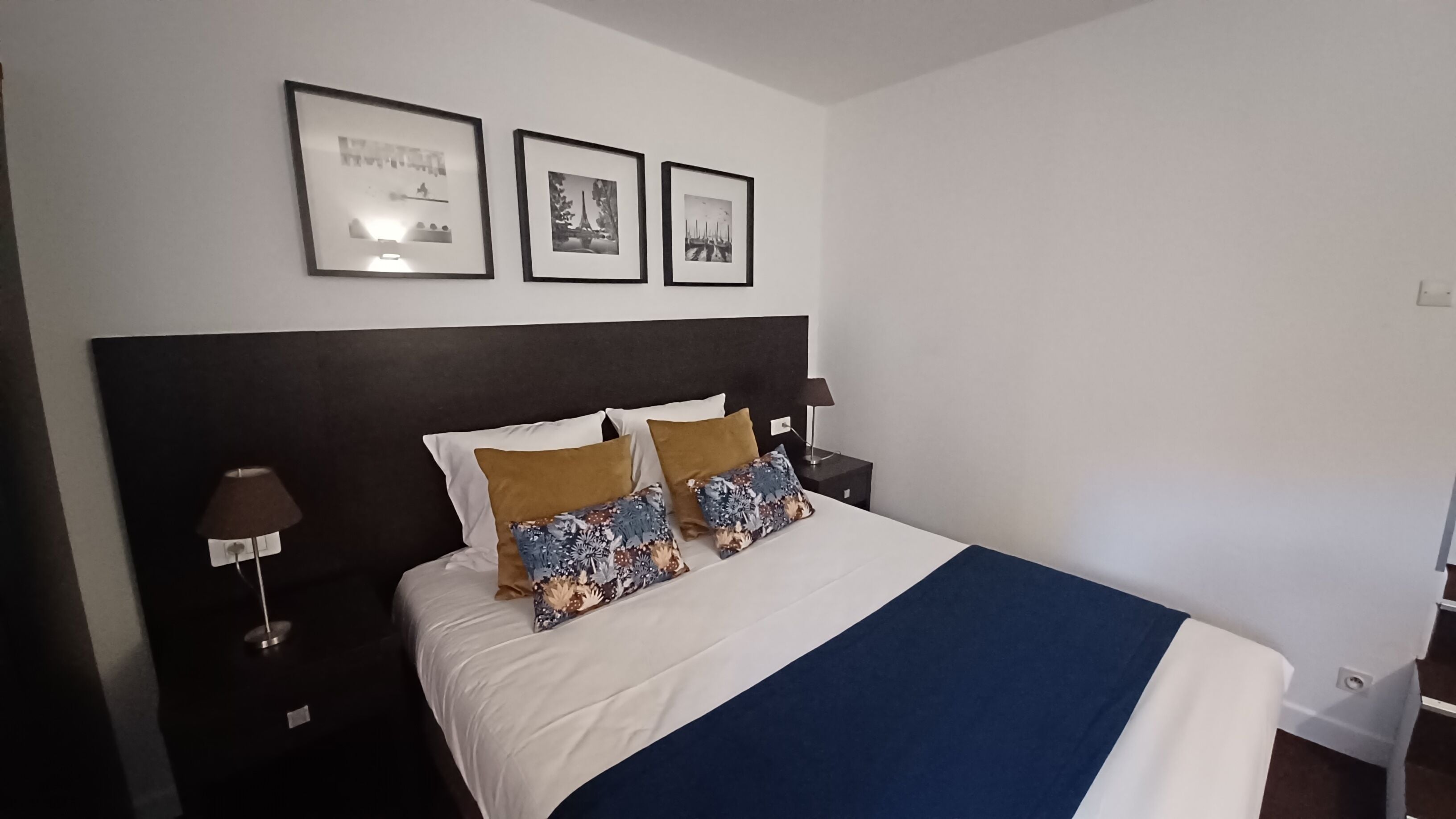 Cama bem arrumada com colcha branca e azul, duas almofadas marrons e uma almofada floral. Há duas mesas de cabeceira com abajures, e três quadros na parede acima da cama.