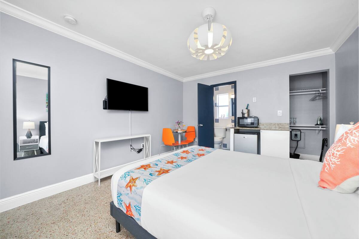 Quarto de hotel moderno com cama, escrivaninha, cadeira, televisão e cozinha compacta.