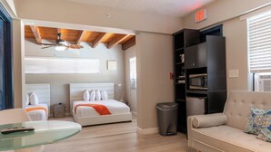 Quarto de hotel moderno com cama, sofá, mesa de vidro, lixeira e cozinha compacta.