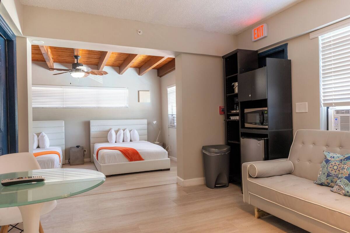 Quarto de hotel moderno com cama, sofá, mesa de vidro, lixeira e cozinha compacta.