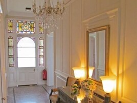 Um corredor com lustre, janelas de vitrais, extintor de incêndio e uma mesa com flores e luminárias.