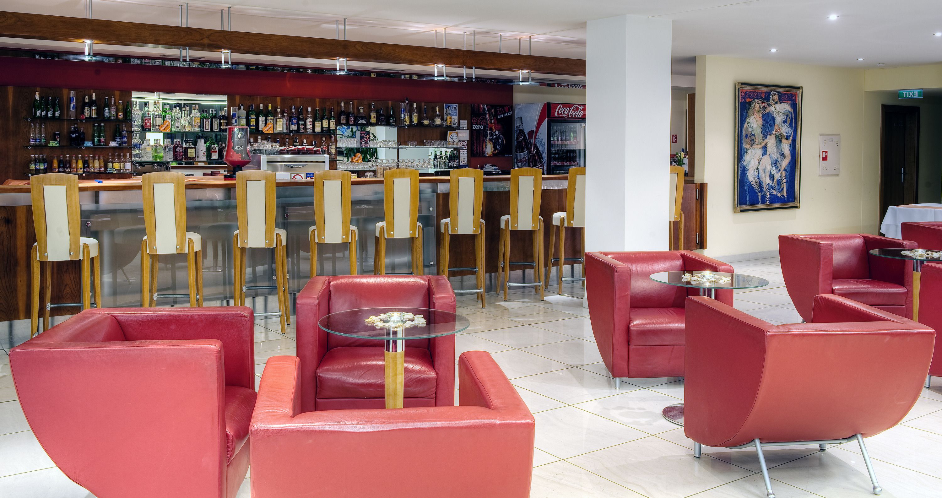 Uma área de bar com diversas opções de assento, incluindo cadeiras e mesas vermelhas, variedade de garrafas de bebidas alcoólicas em prateleiras e uma grande geladeira Coca-Cola.