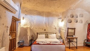Quarto com parede de pedra, chão de madeira, cama com roupa de cama branca, guarda-roupa de madeira, mesa de cabeceira com abajur e espelho na parede.