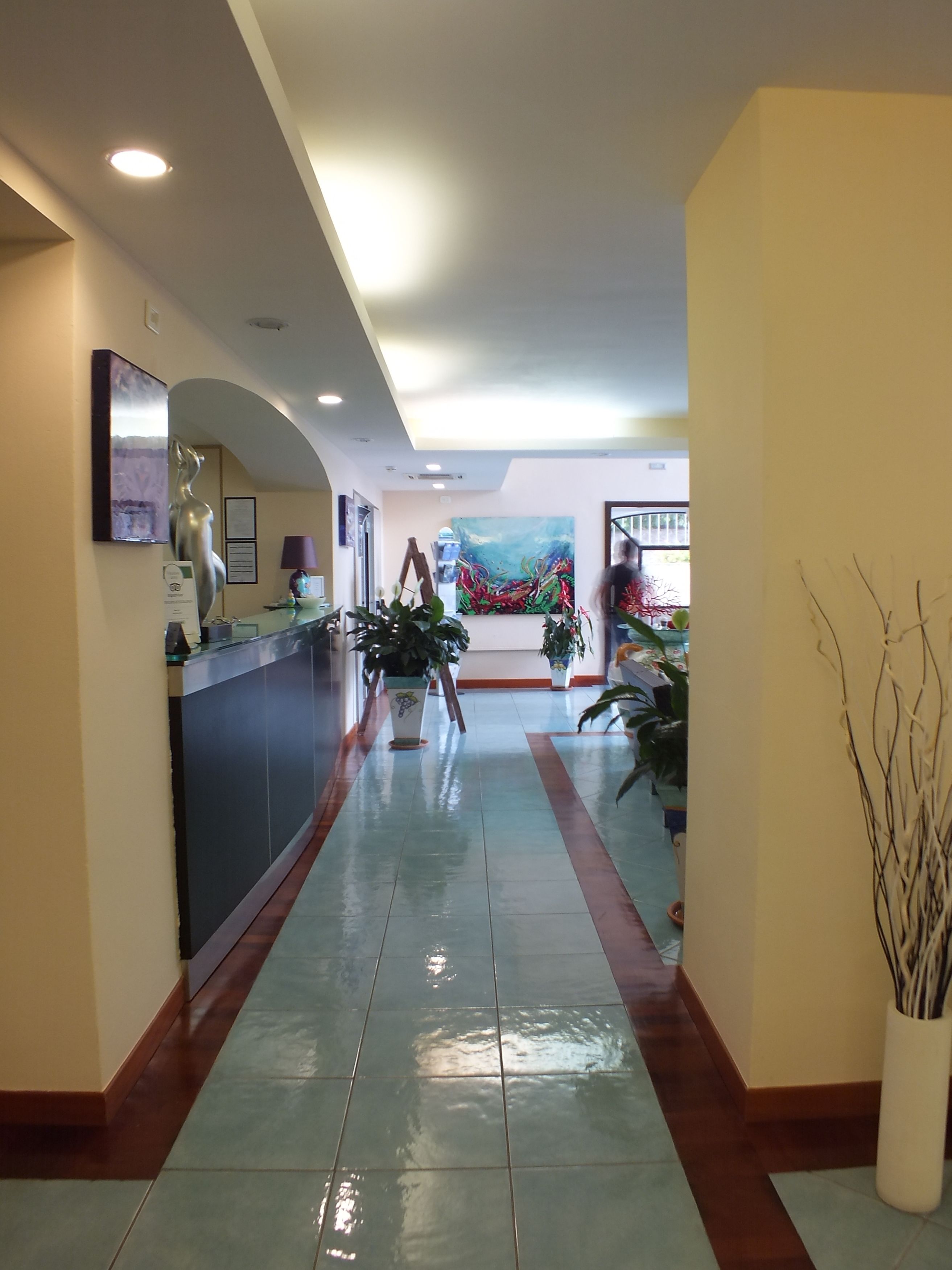 Uma área de recepção de hotel com piso de azulejos, balcão de recepção, um quadro na parede e plantas em vasos.