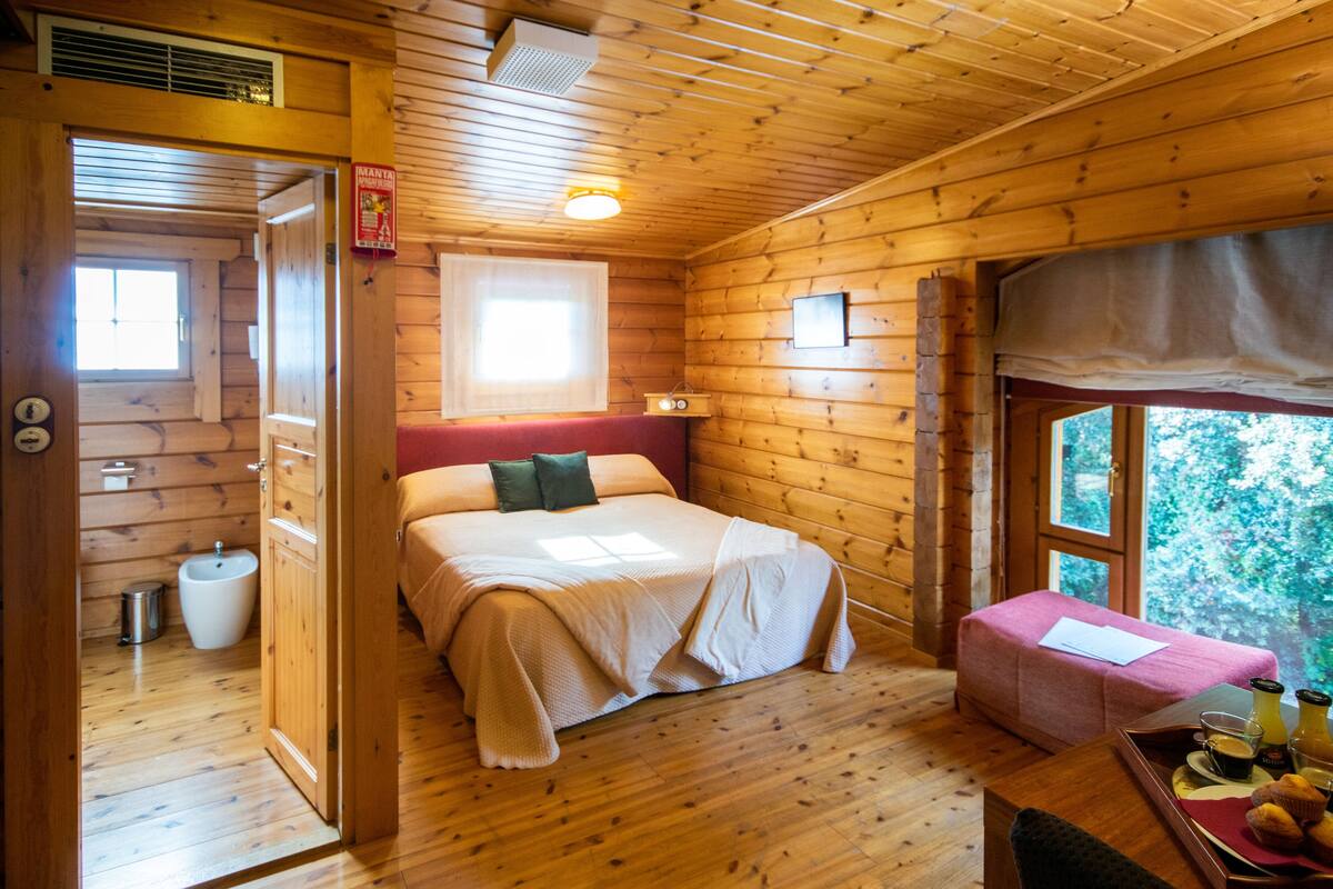 Quarto de cabana de madeira com uma cama, uma mesinha com itens de café da manhã, uma janela com vista para árvores e uma porta que dá para um banheiro.