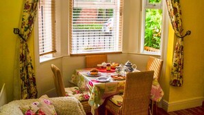 Uma área de refeições com uma mesa posta para dois, rodeada por cadeiras de vime, com uma janela que oferece vista para o verde lá fora.