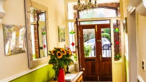 Um corredor com carpete vermelho, uma escrivaninha de madeira com um vaso de girassóis, um candelabro e uma porta que dá para o exterior.