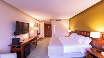 Quarto de hotel com cama, escrivaninha, cadeira, televisão e luminárias.
