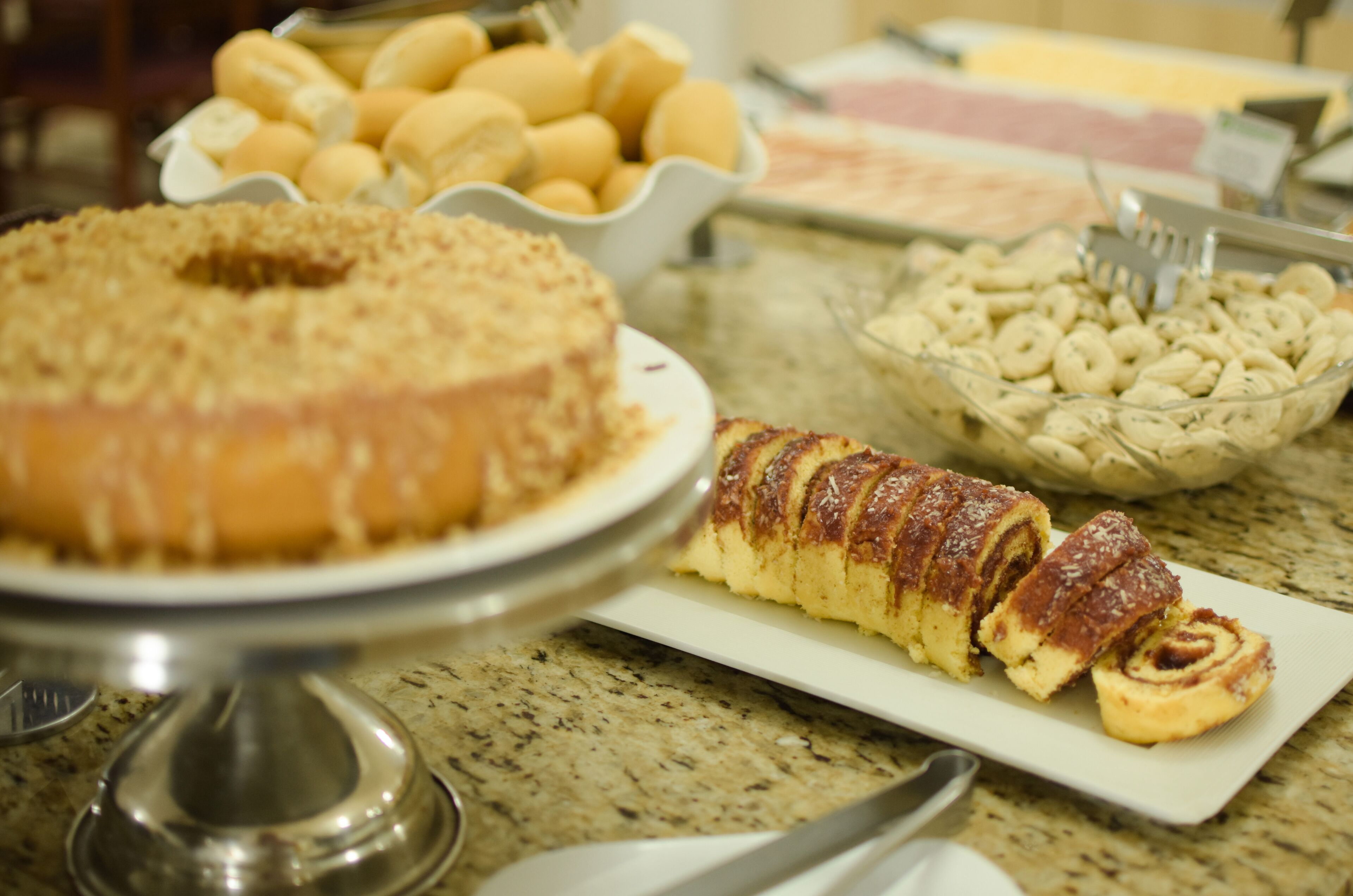 Buffet com diversos produtos de panificação, incluindo um bolo bundt, um pastel enrolado e uma tigela de cereais.