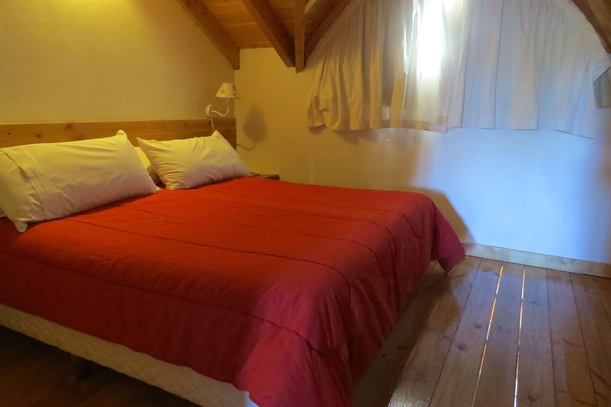 Quarto com cama, vigas de madeira no teto, um ventilador fixado na parede e uma janela com cortinas.
